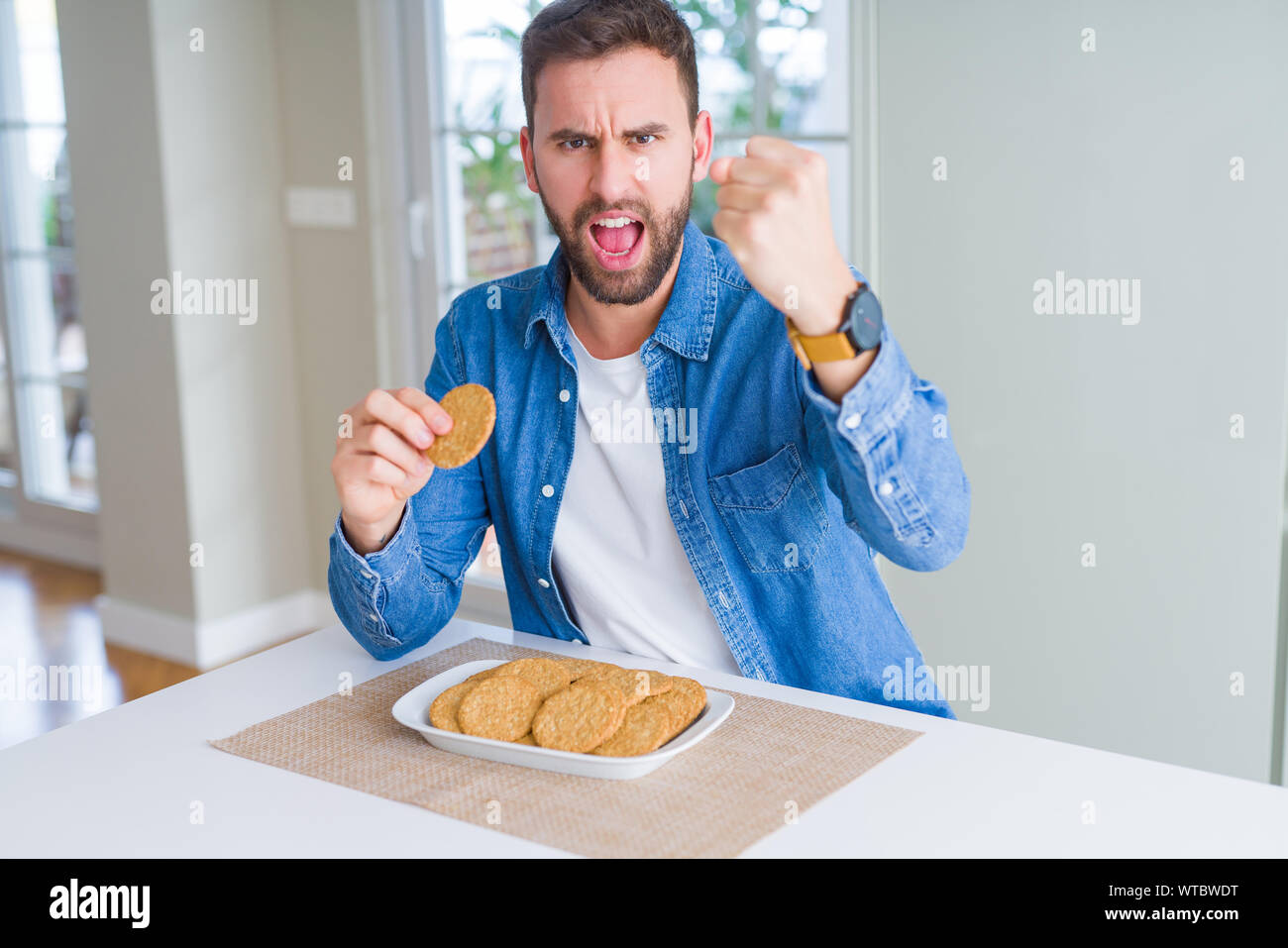 Handsome man eating healthy whole grain biscuit annoyed and frustrated ...