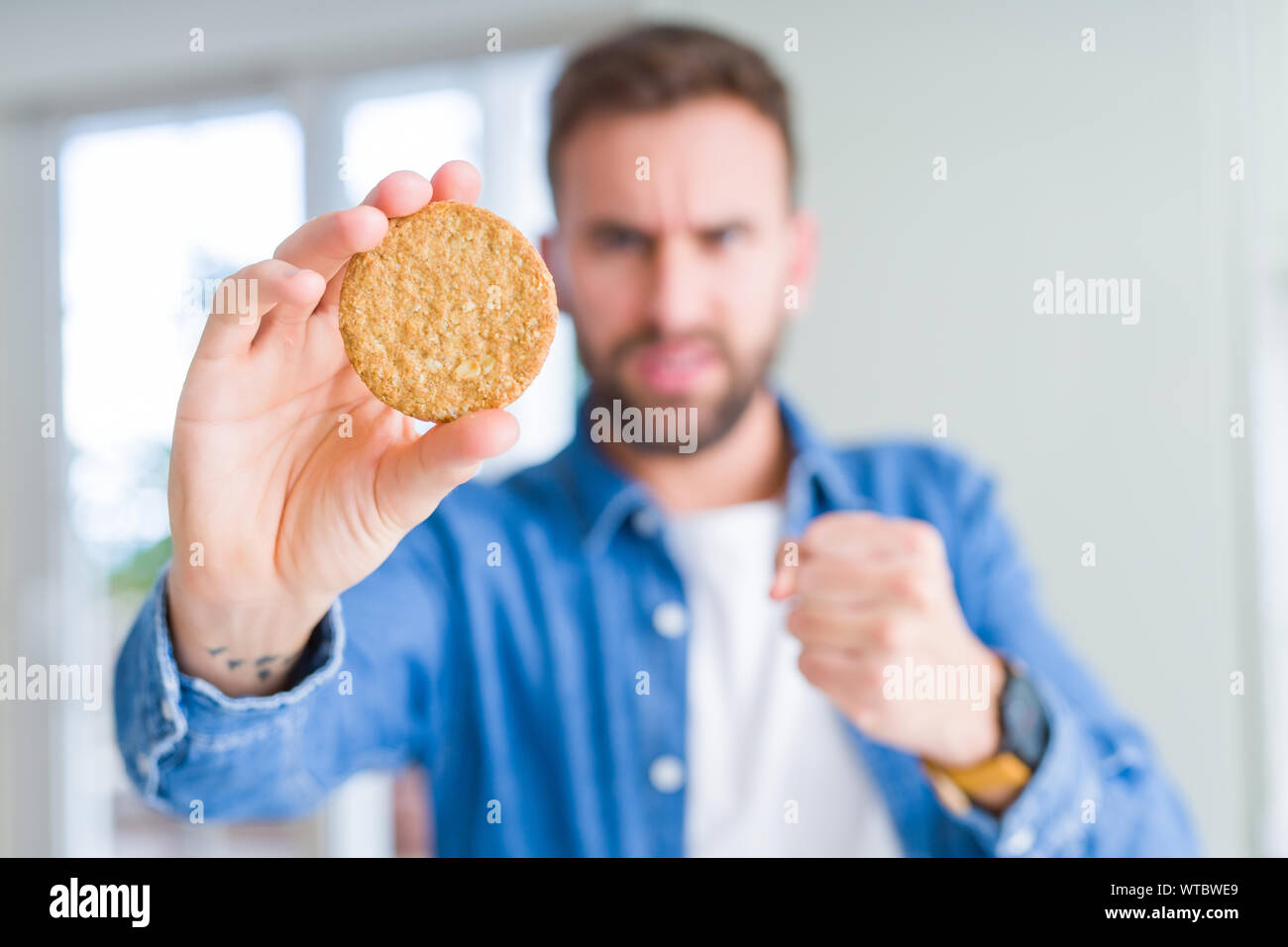 Handsome man eating healthy whole grain biscuit annoyed and frustrated ...