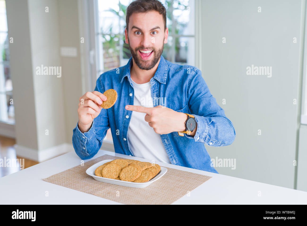 Handsome man eating healthy whole grain biscuit very happy pointing ...