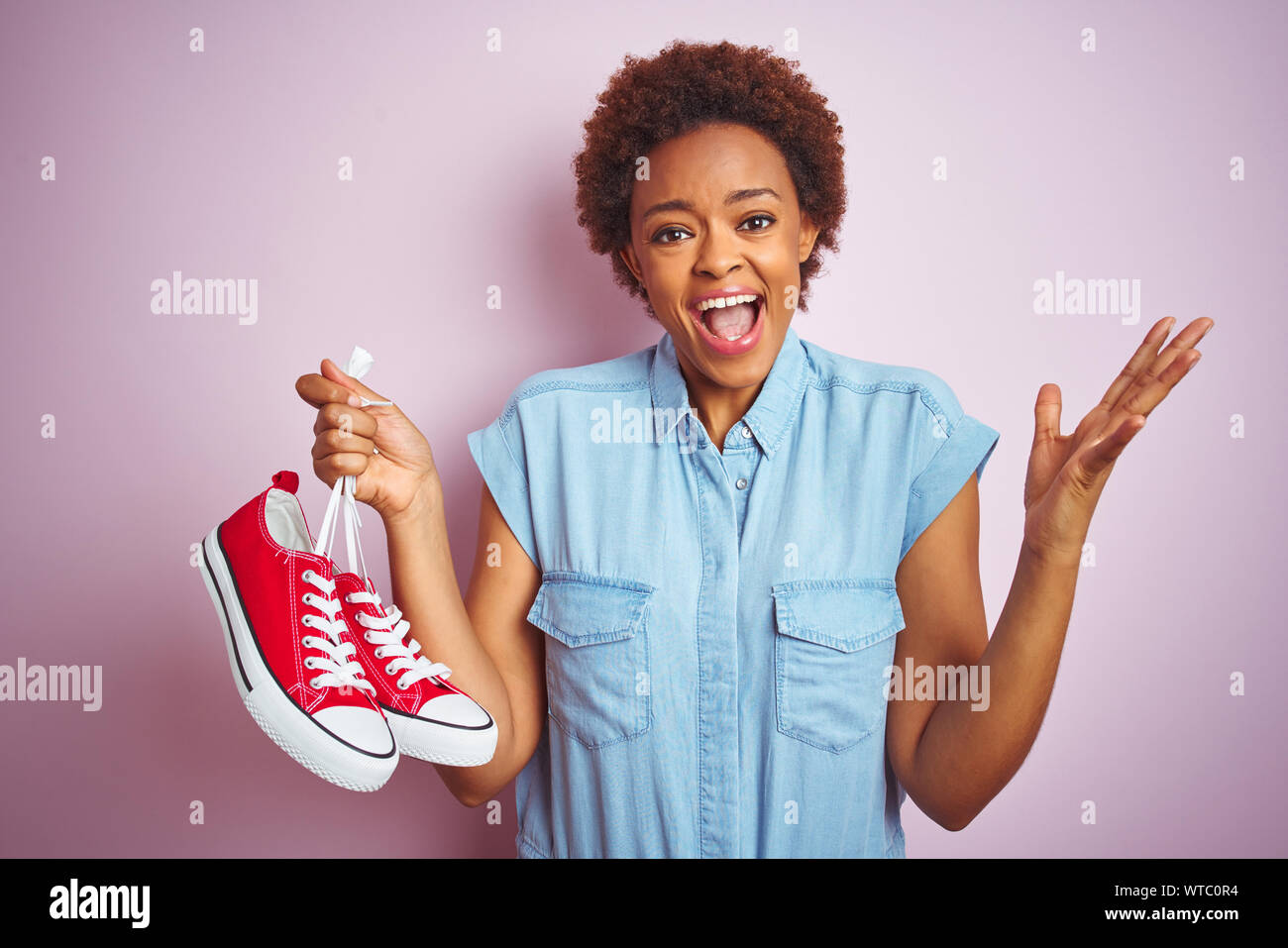 African american woman holding red casual sneakers over pink isolated ...