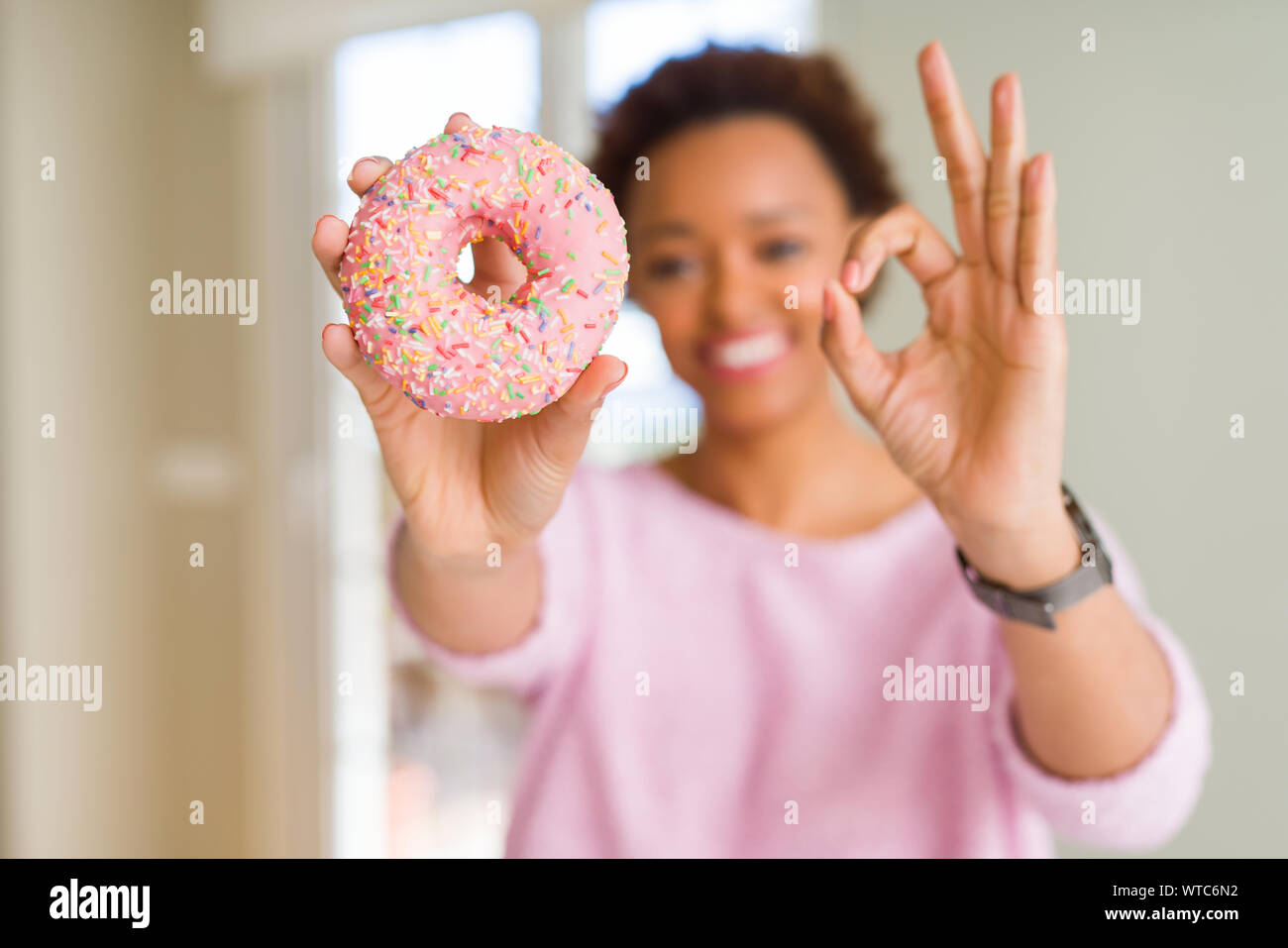 Young african american woman eating pink sugar donut doing ok sign with ...