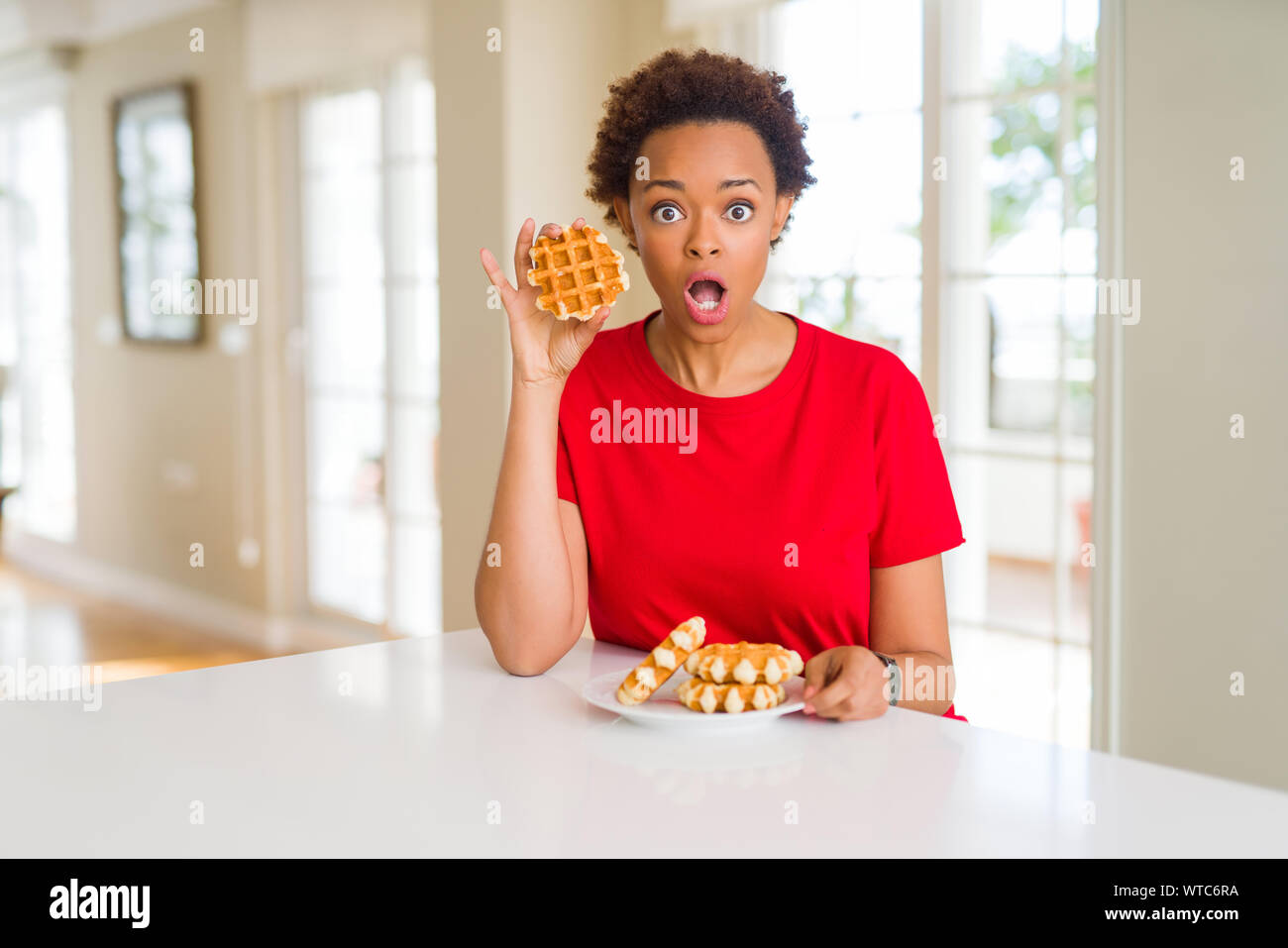 Young african american woman eating sweet waffle scared in shock with a ...