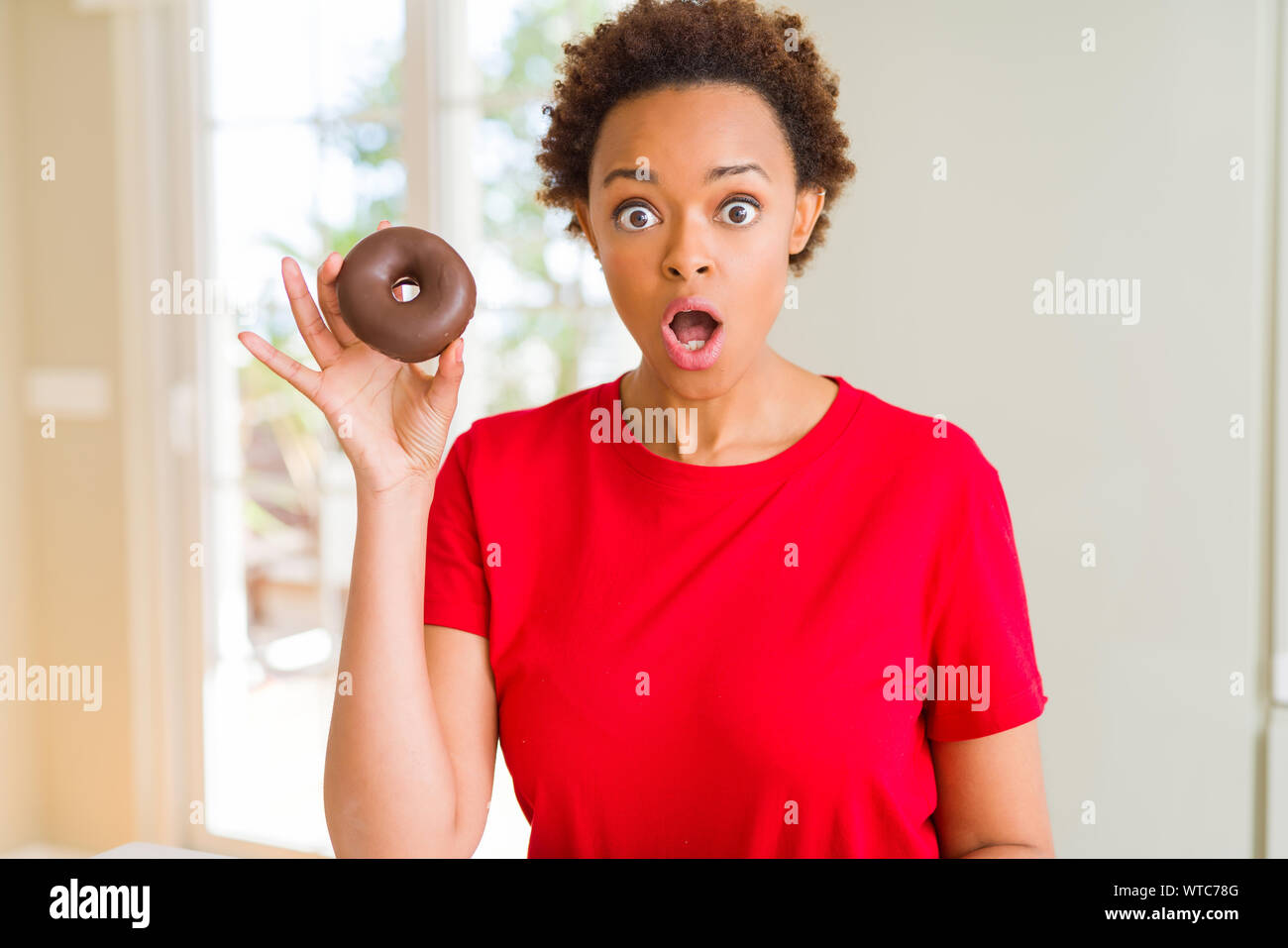 Young african american woman eating chocolate donut scared in shock ...