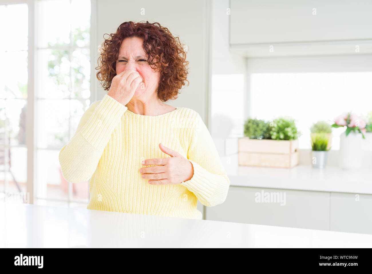 Beautiful senior woman wearing yellow sweater smelling something stinky ...