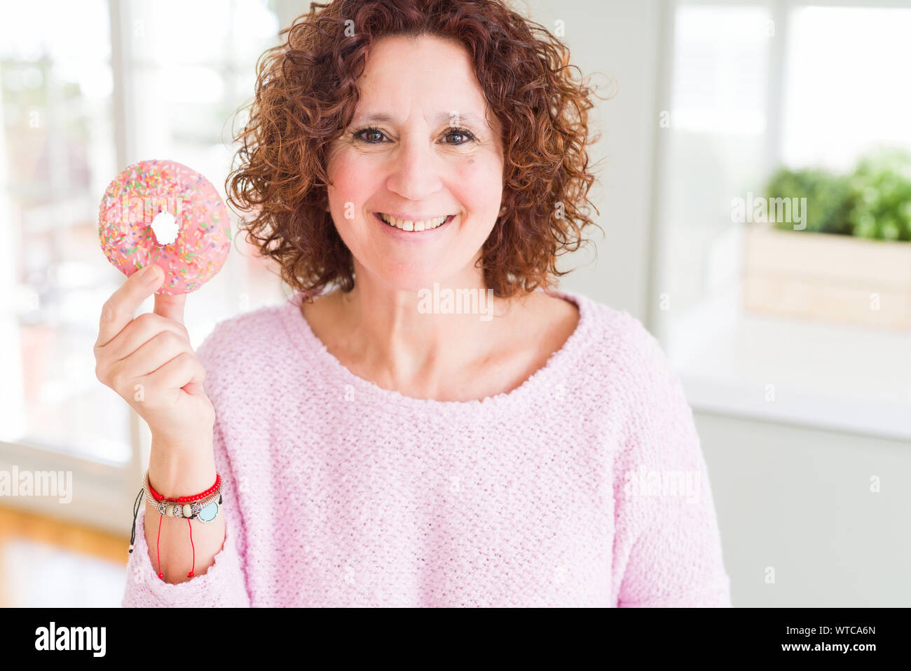 Senior woman eating pink sugar donut with a happy face standing and ...