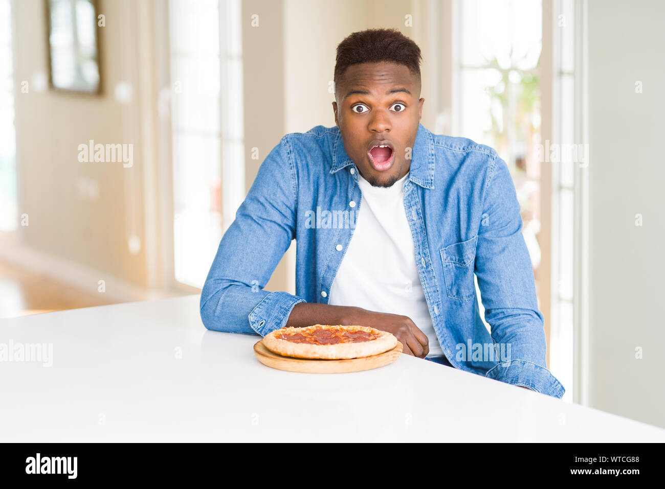African american man eating pepperoni pizza at home scared in shock ...