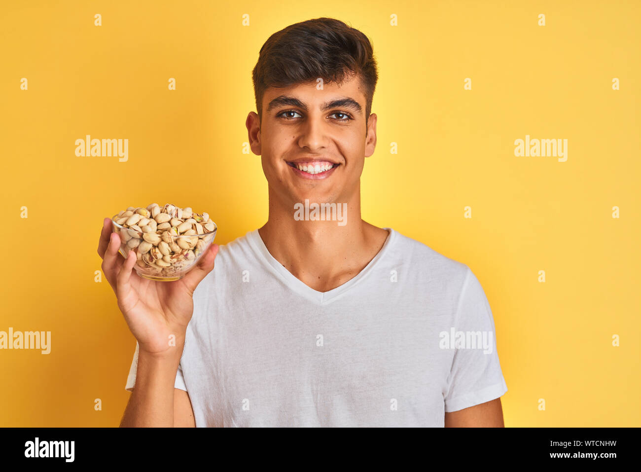 Young indian man holding bowl with pistachios over isolated yellow ...