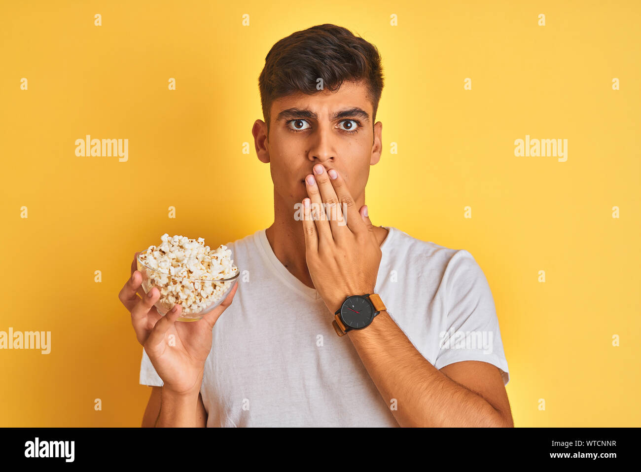 Young indian man holding pack of popcorn standing over isolated yellow ...