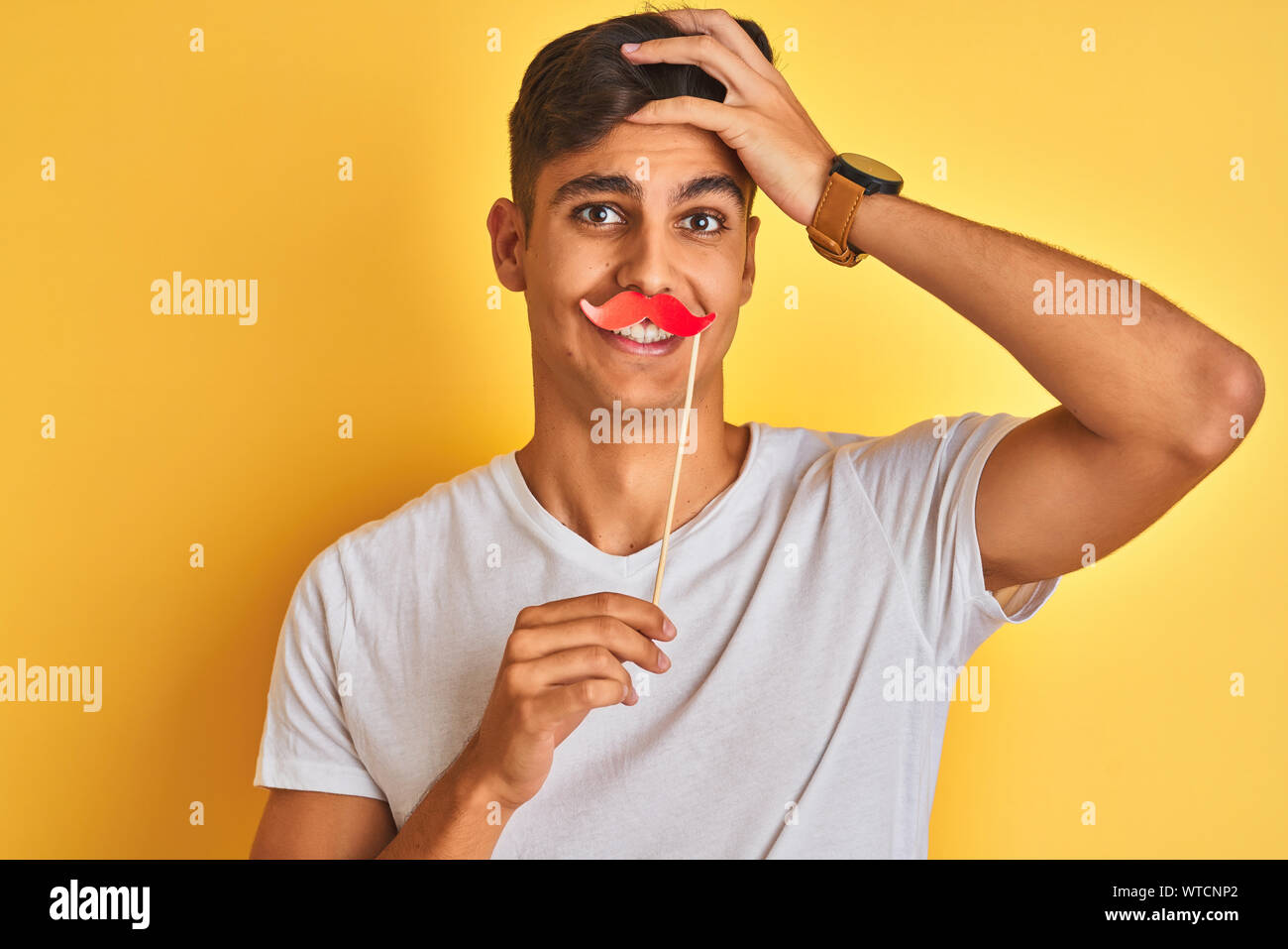 Young indian man holding fanny mustache standing over isolated yellow ...