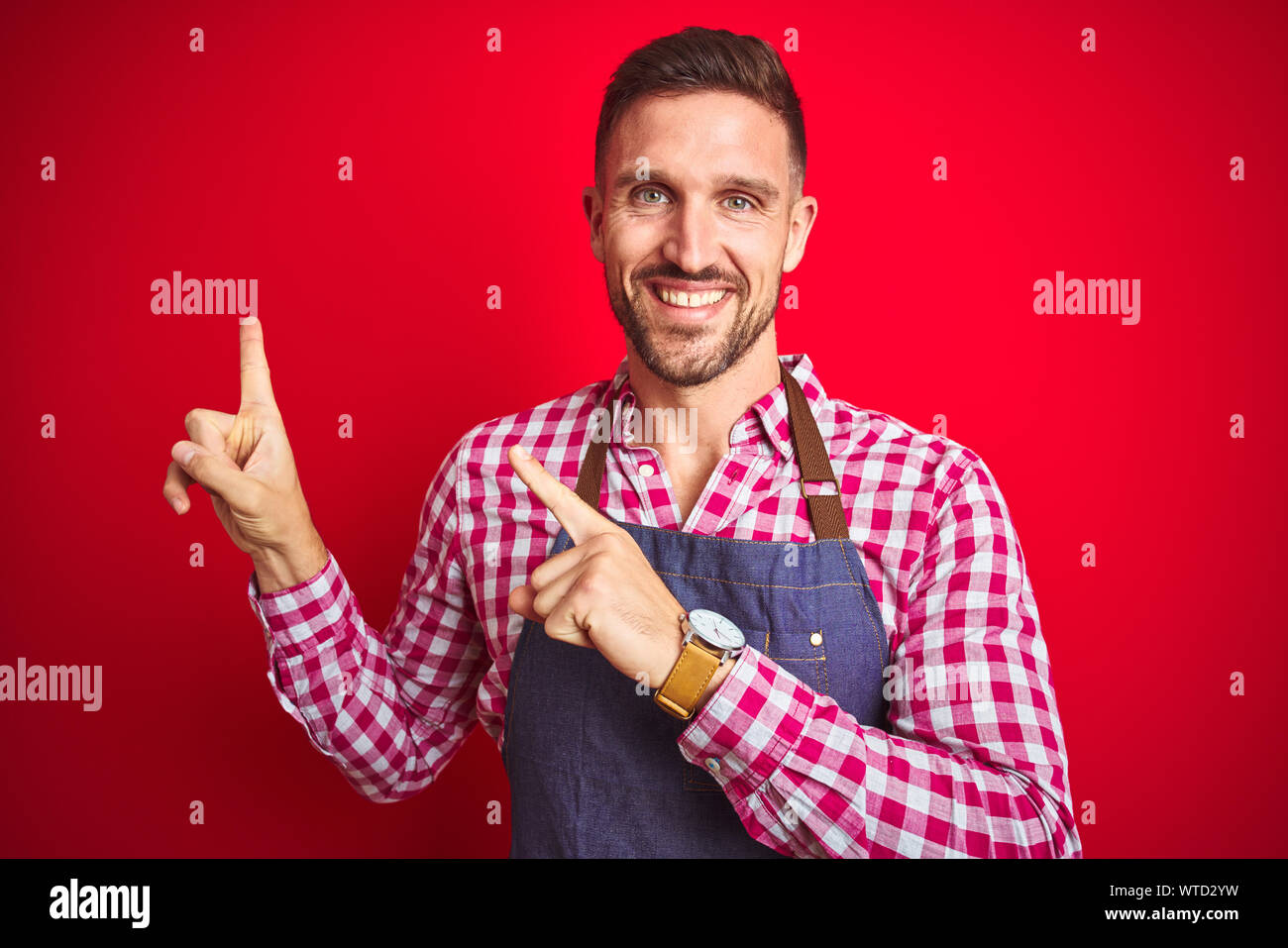 Young handsome man wearing shop owner apron uniform over red isolated ...