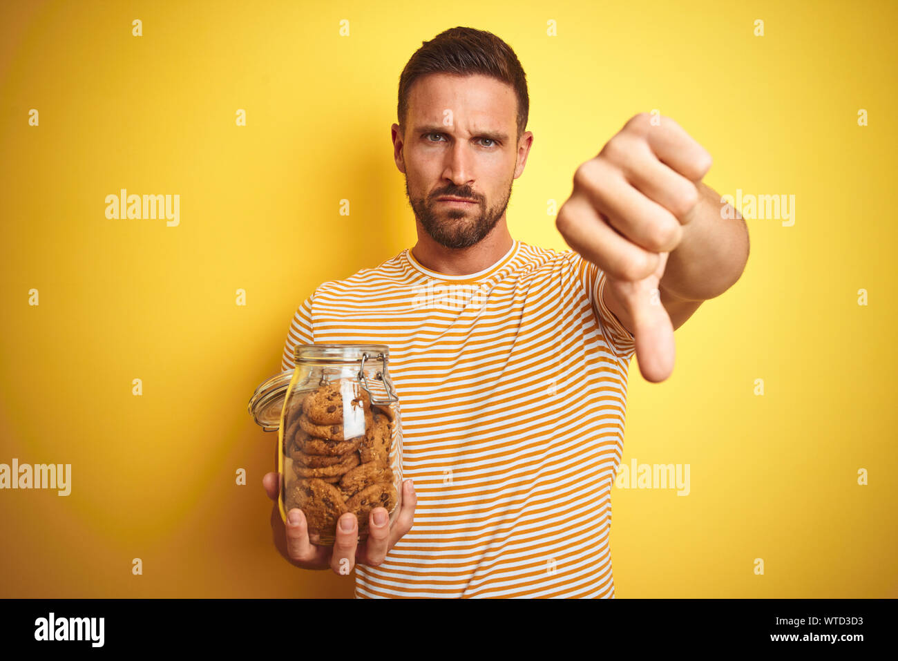 Young handsome man holding a jar of cookies over yellow isolated ...