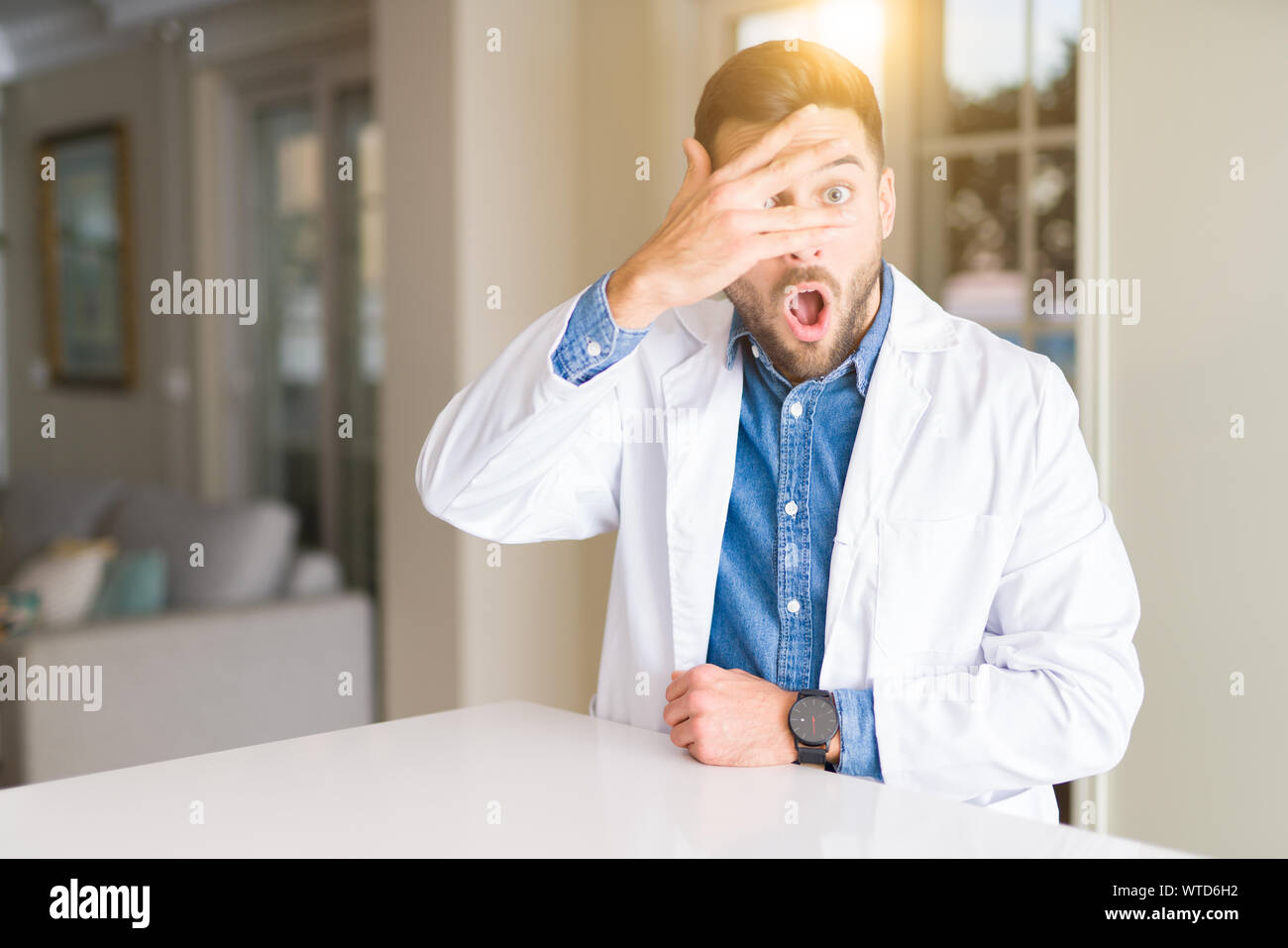 Young handsome doctor man at the clinic peeking in shock covering face and eyes with hand ...