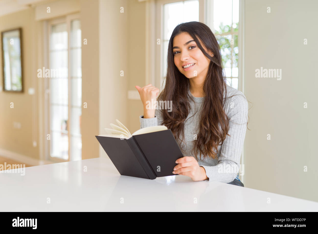 Young woman reading a book pointing and showing with thumb up to the ...