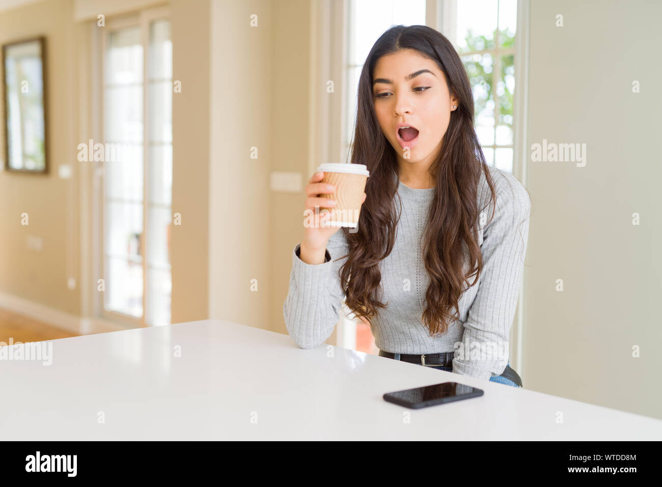 Young woman drinking a cup of coffee at home scared in shock with a ...