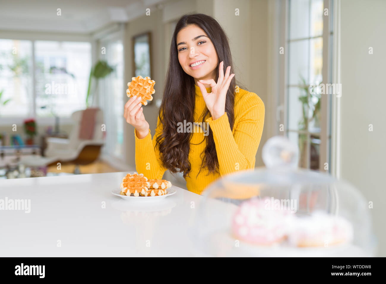 Young woman eating sweet waffle pastry doing ok sign with fingers ...