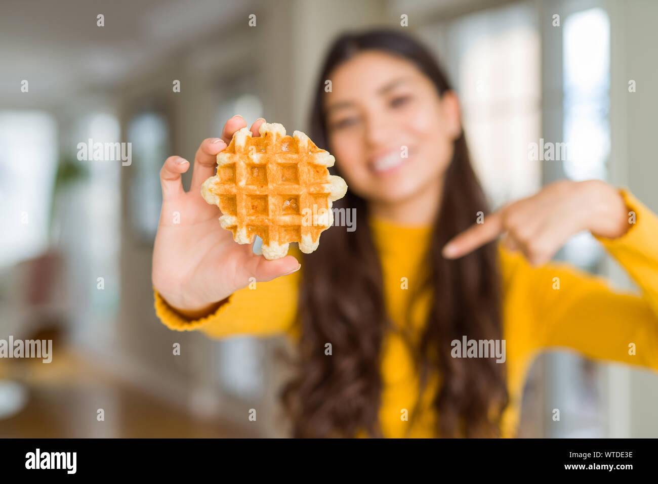 Young woman eating sweet waffle pastry with surprise face pointing ...
