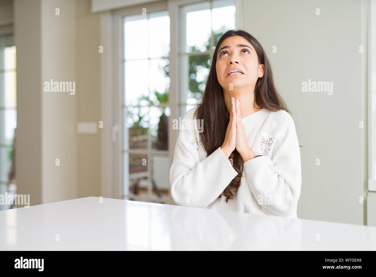 Young beautiful woman at home on white table begging and praying with ...