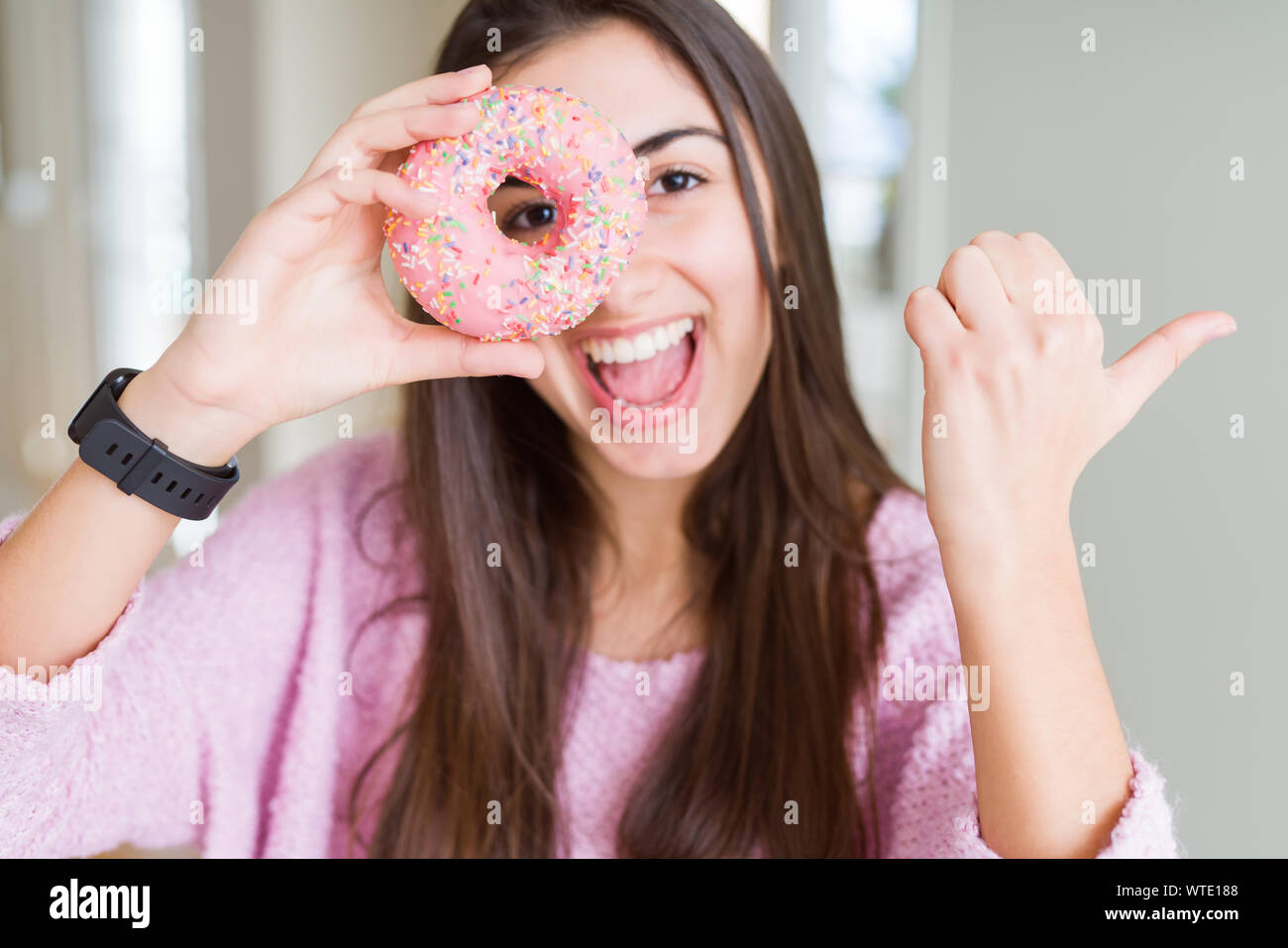 Beautiful young woman eating pink chocolate chips donut pointing and ...