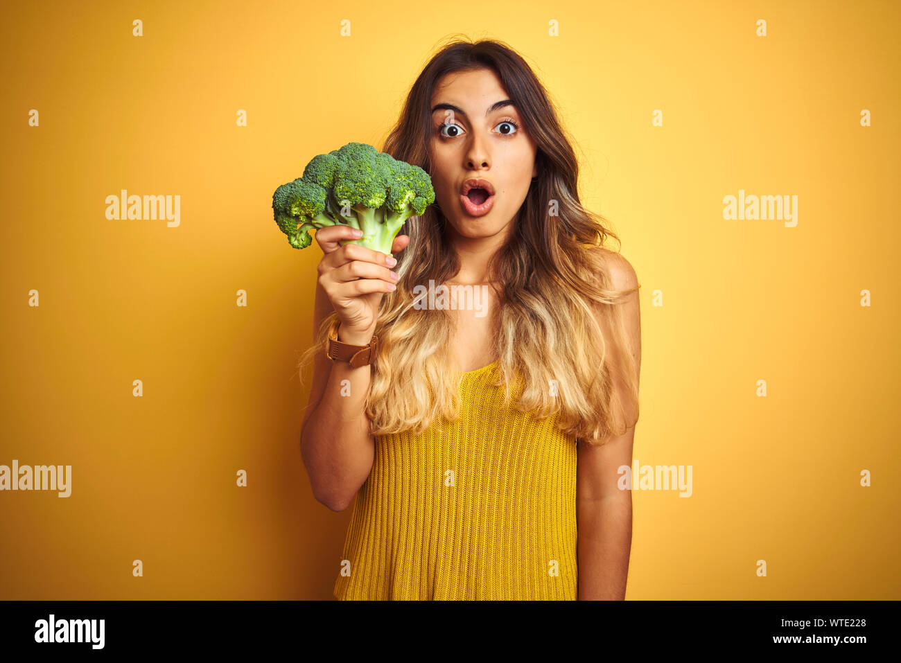 Young beautiful woman eating broccoli over yellow isolated background ...