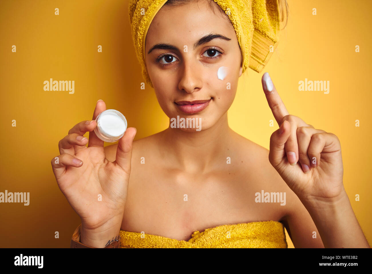 Young woman wearing a shower towel using face cream over yellow ...