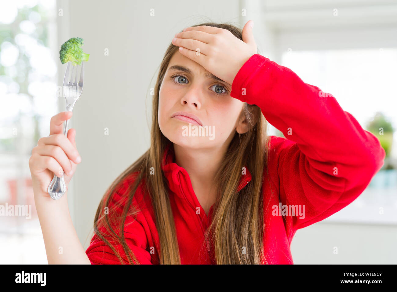 Beautiful young girl eating fresh broccoli stressed with hand on head ...