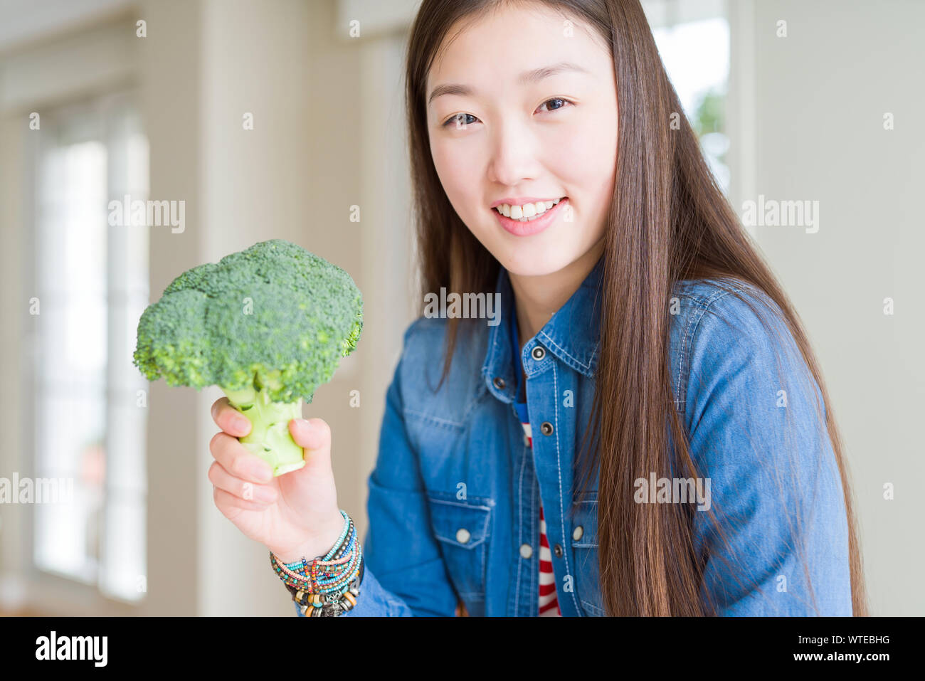 Beautiful Asian woman eating green fresh broccoli with a happy face standing and smiling with a ...