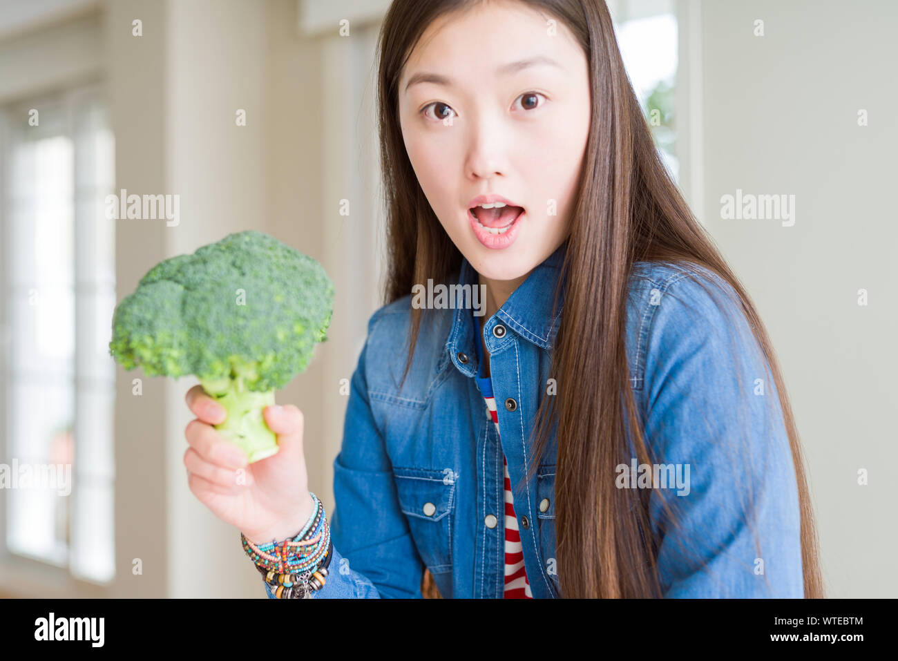Beautiful Asian woman eating green fresh broccoli scared in shock with ...