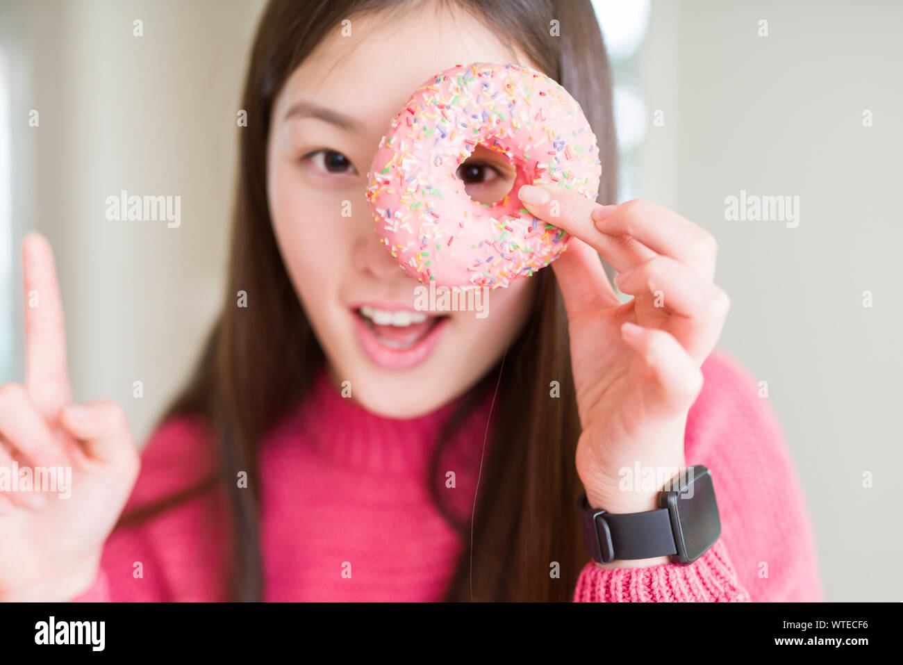 Beautiful Asian woman eating pink sugar donut surprised with an idea or ...