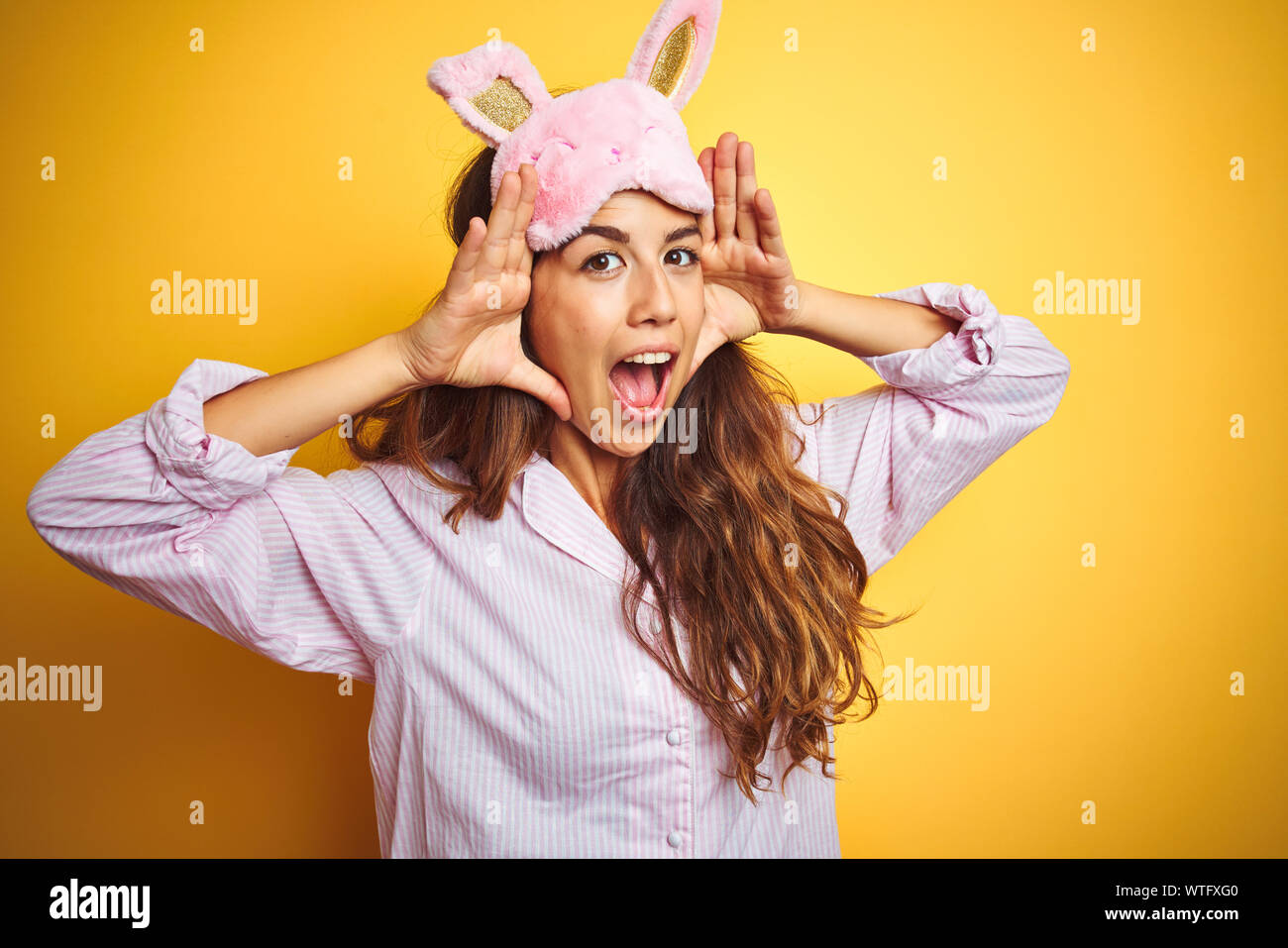 Young woman wearing pajama and sleep mask standing over yellow isolated ...