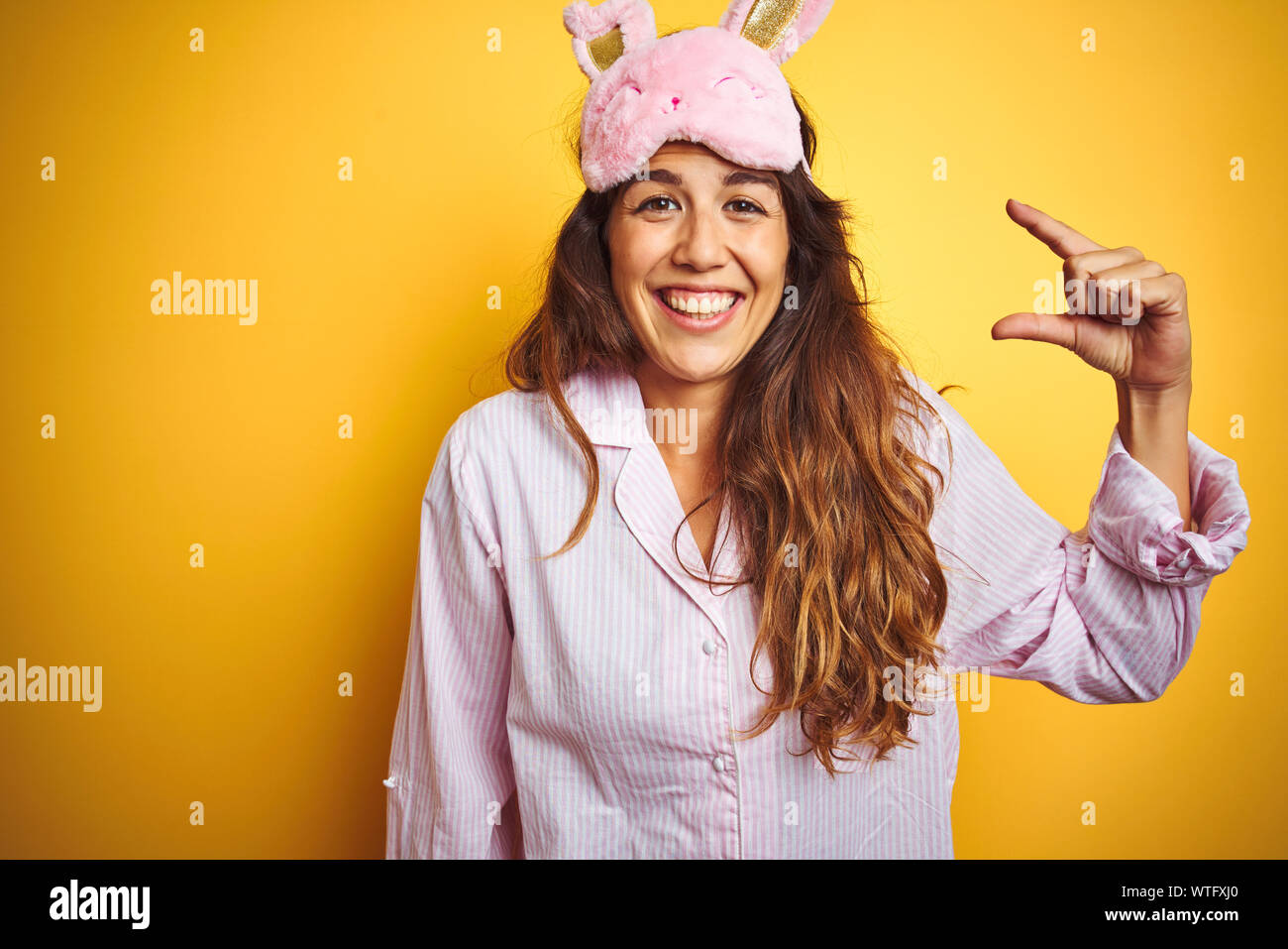 Young woman wearing pajama and sleep mask standing over yellow isolated ...