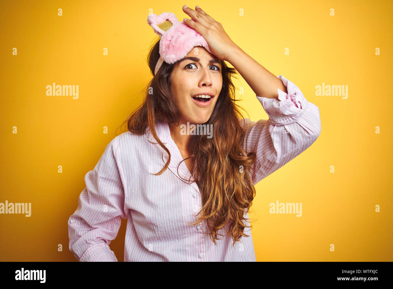 Young woman wearing pajama and sleep mask standing over yellow isolated ...