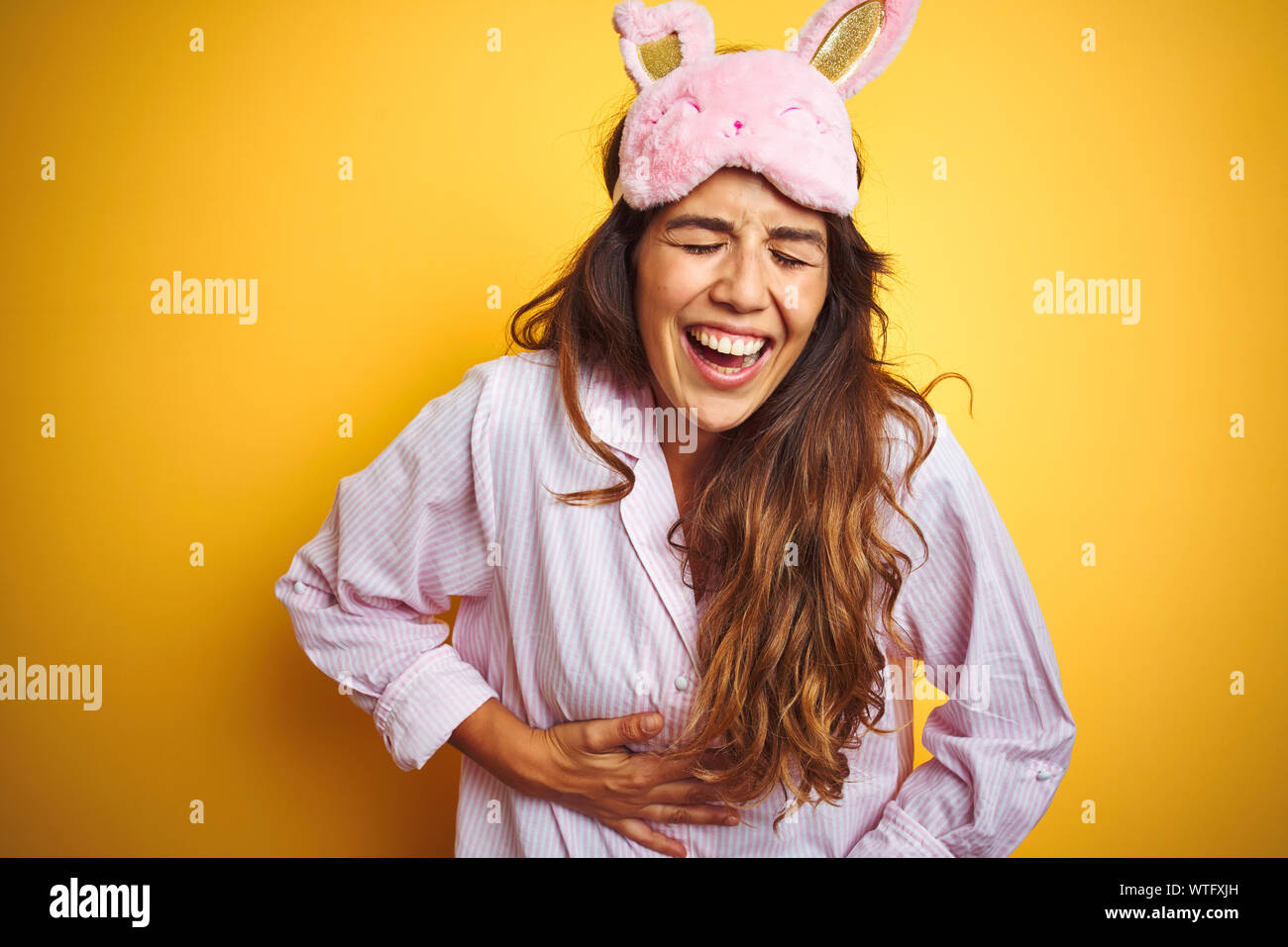 Young woman wearing pajama and sleep mask standing over yellow isolated ...