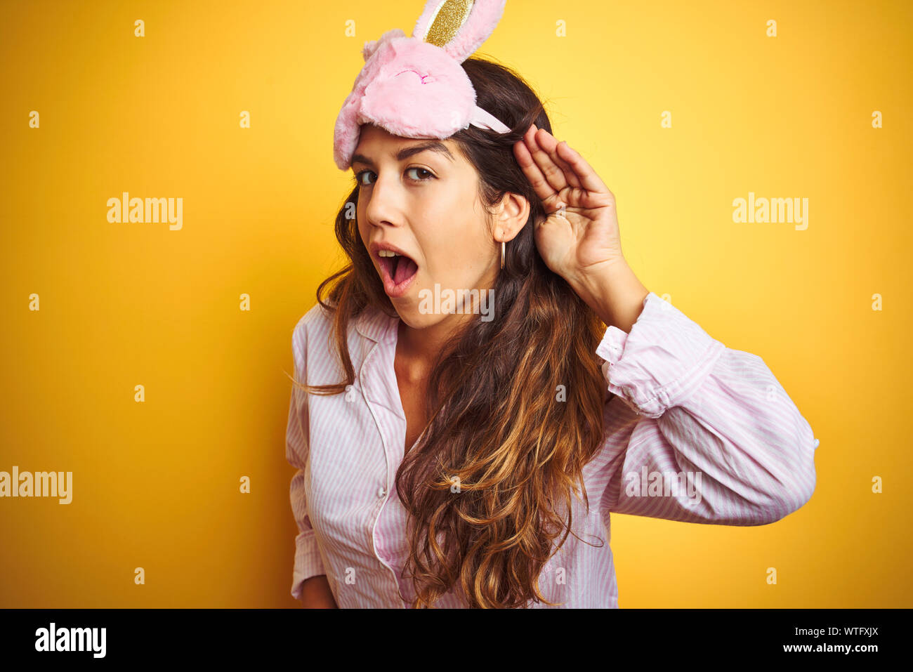 Young woman wearing pajama and sleep mask standing over yellow isolated ...