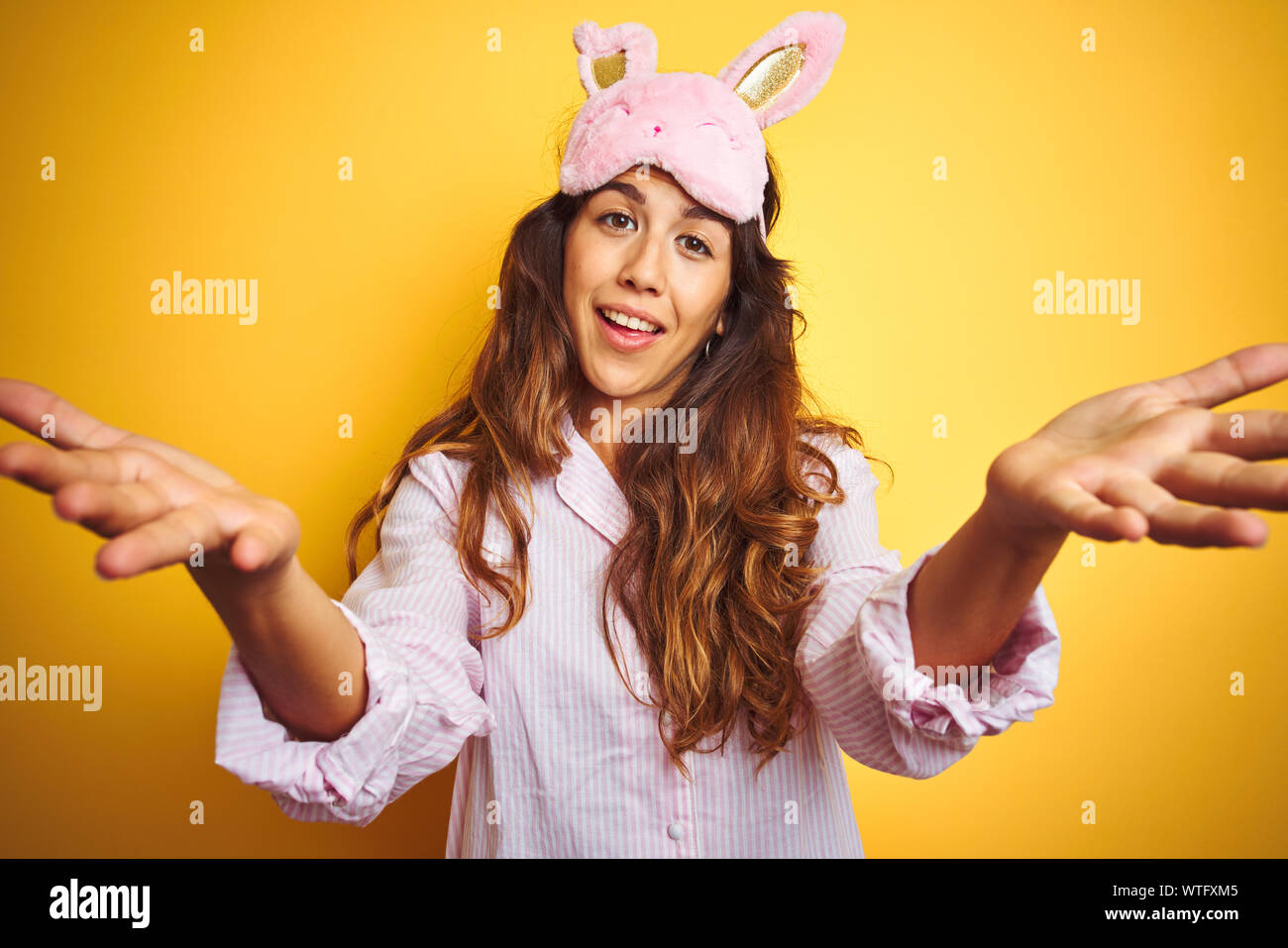 Young woman wearing pajama and sleep mask standing over yellow isolated ...