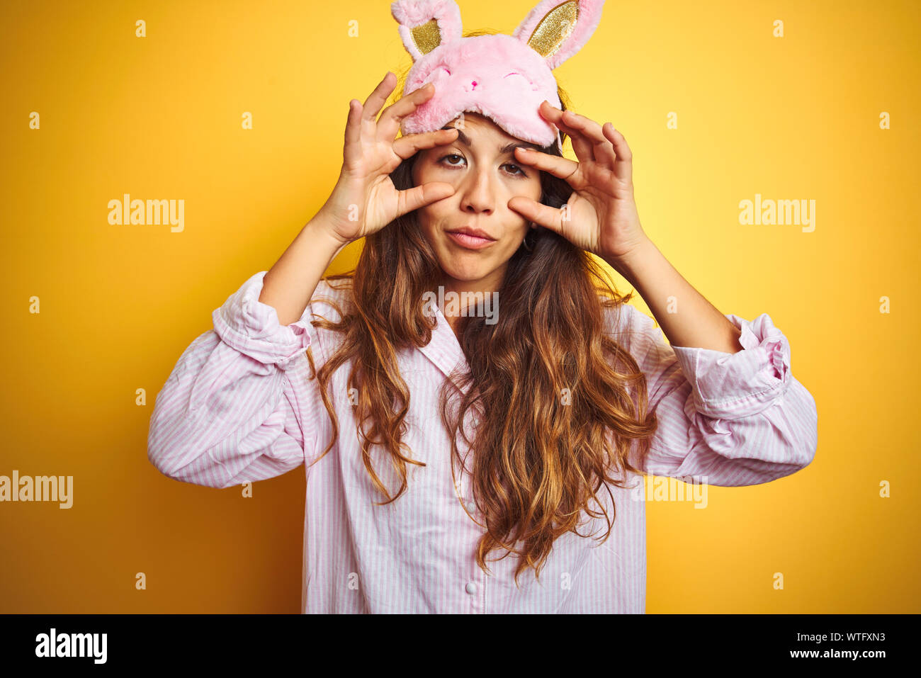 Young woman wearing pajama and sleep mask standing over yellow isolated ...