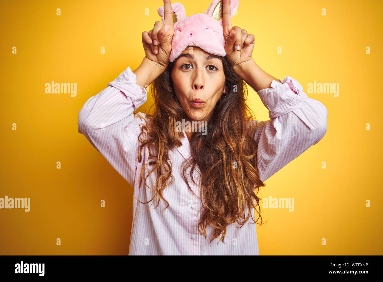 Young woman wearing pajama and sleep mask standing over yellow isolated ...