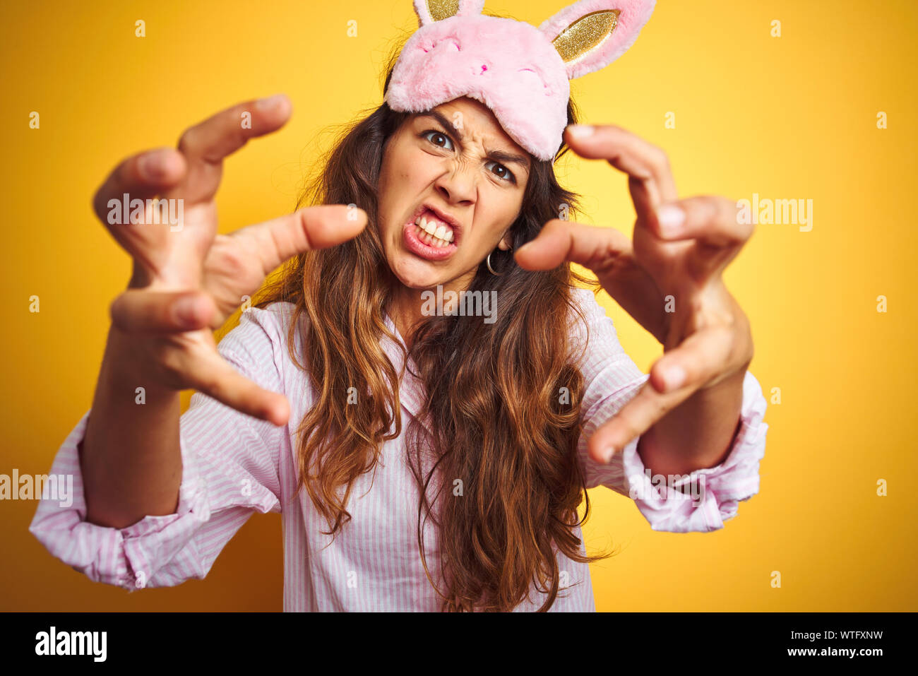 Young woman wearing pajama and sleep mask standing over yellow isolated ...