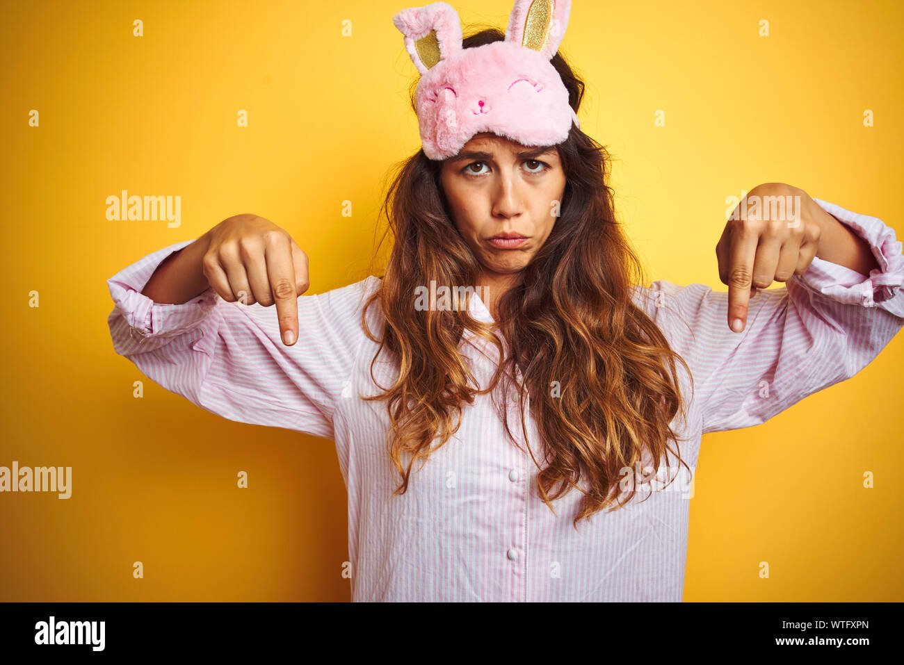 Young woman wearing pajama and sleep mask standing over yellow isolated ...