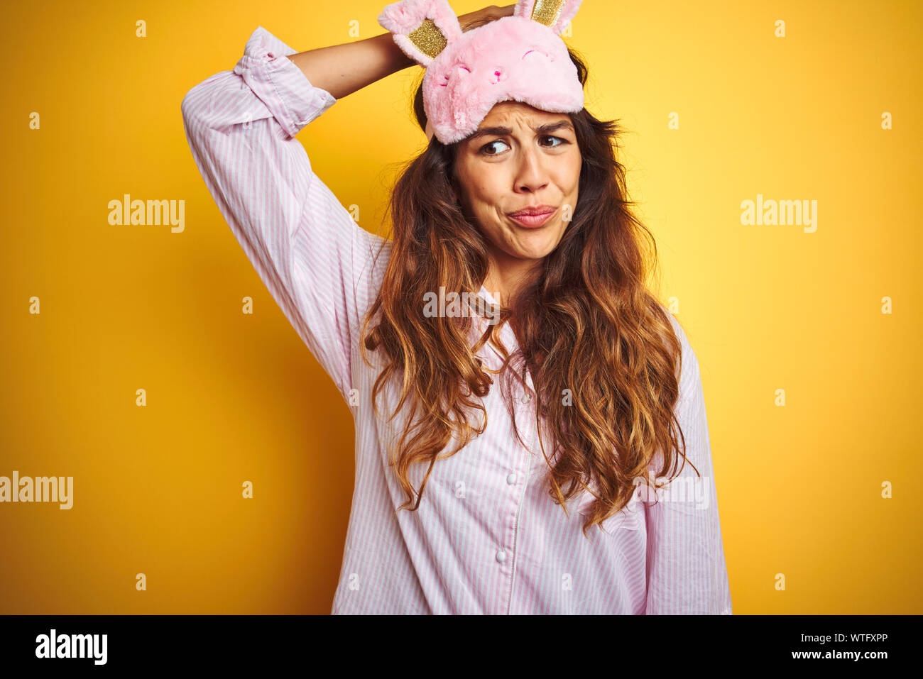Young woman wearing pajama and sleep mask standing over yellow isolated ...