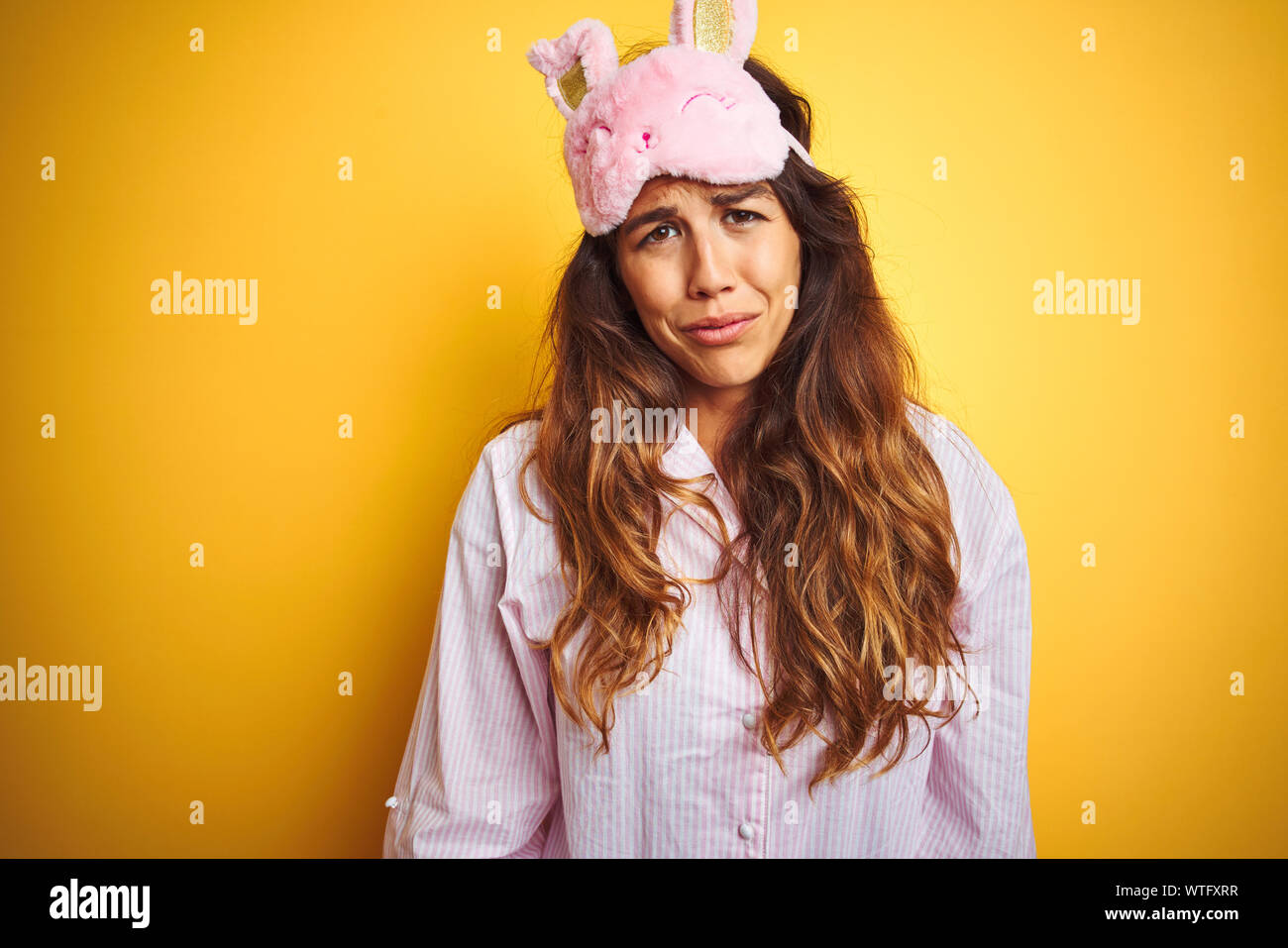 Young woman wearing pajama and sleep mask standing over yellow isolated ...