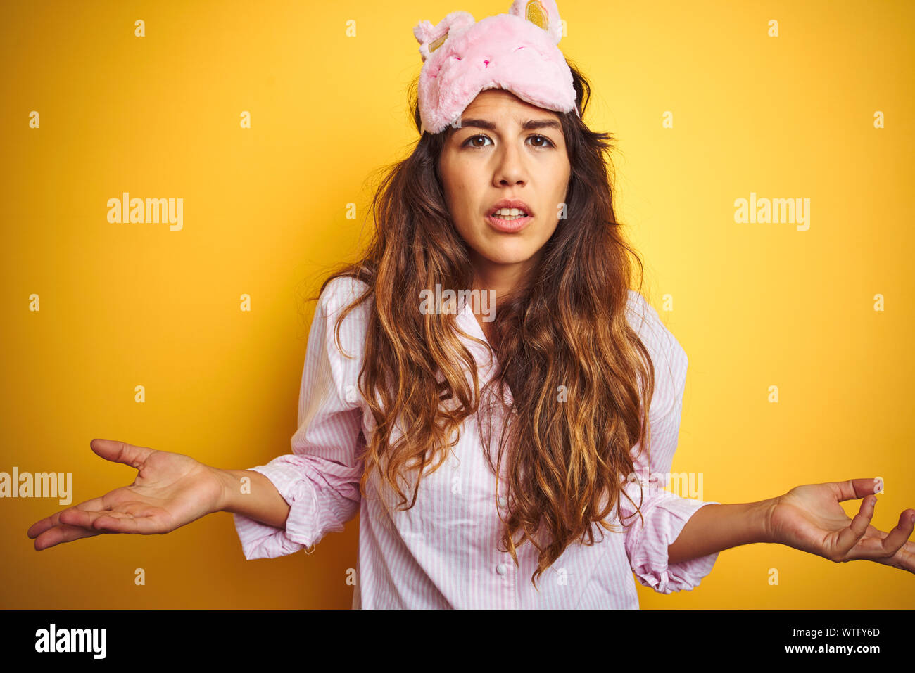 Young woman wearing pajama and sleep mask standing over yellow isolated ...
