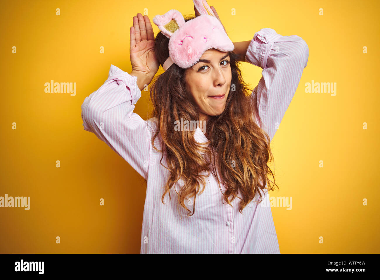 Young woman wearing pajama and sleep mask standing over yellow isolated ...