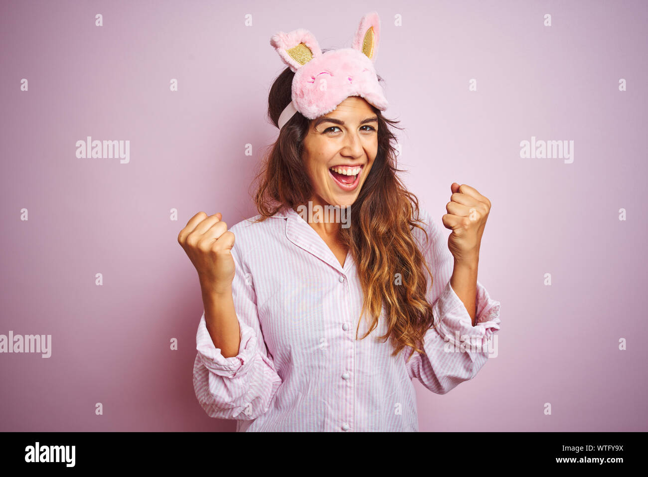 Young woman wearing pajama and sleep mask standing over pink isolated ...