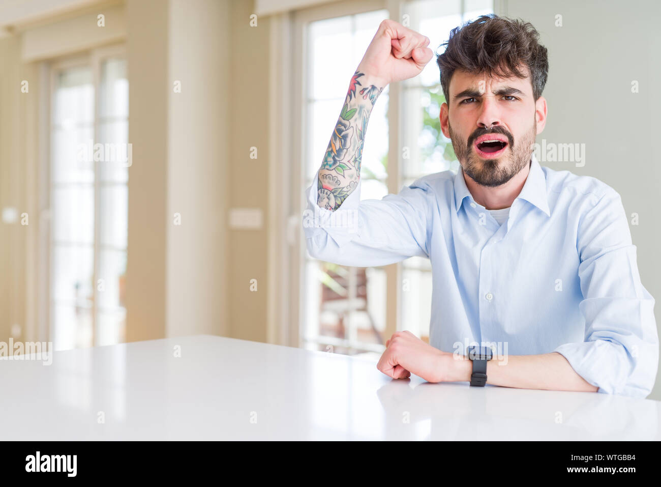 Young businesss man sitting on white table angry and mad raising fist ...