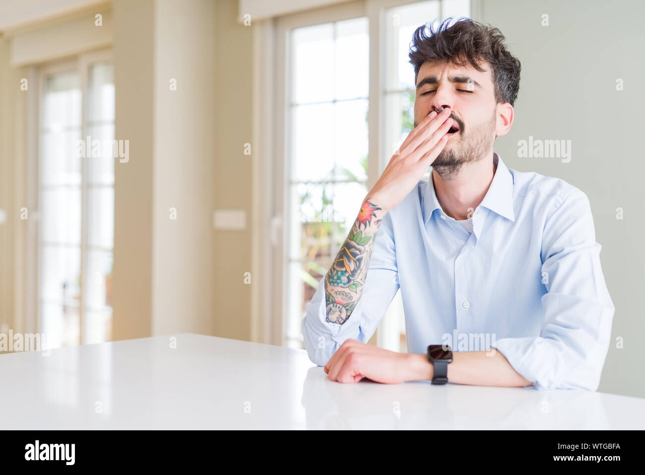 Young businesss man sitting on white table bored yawning tired covering ...