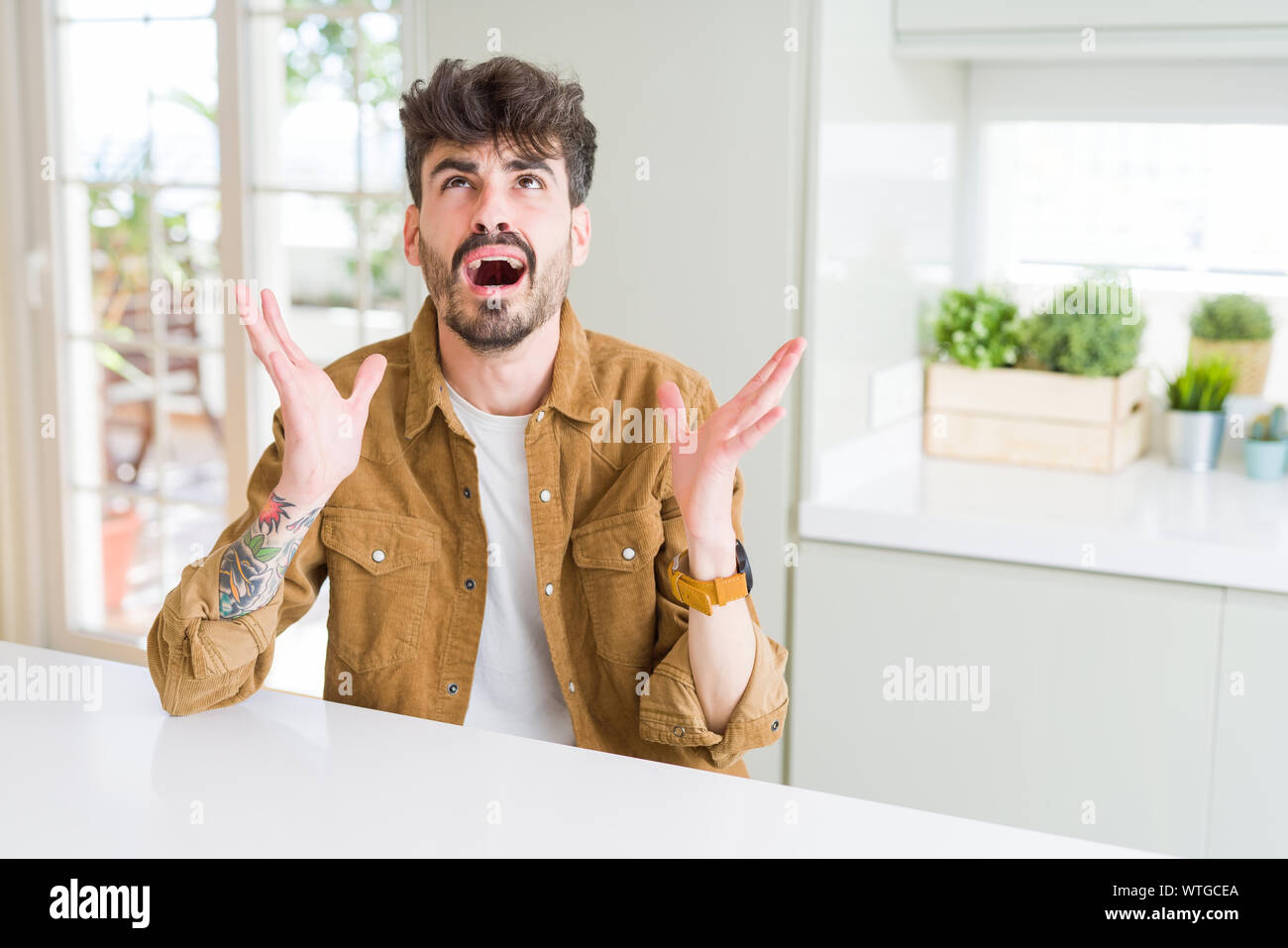 Young man wearing casual jacket sitting on white table crazy and mad ...