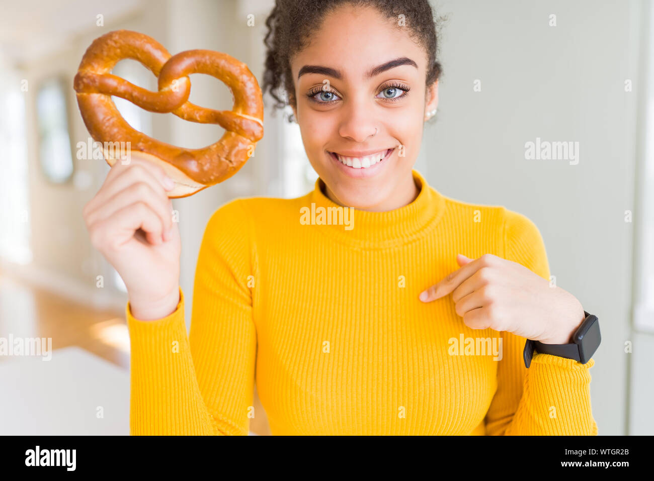 Young african american woman eating German salty pastry pretzel with surprise face pointing ...