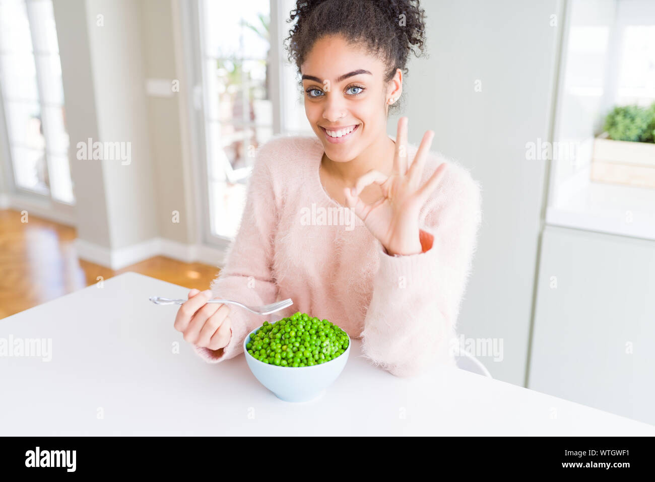 Young african american girl eating healthy green peas doing ok sign with fingers, excellent ...