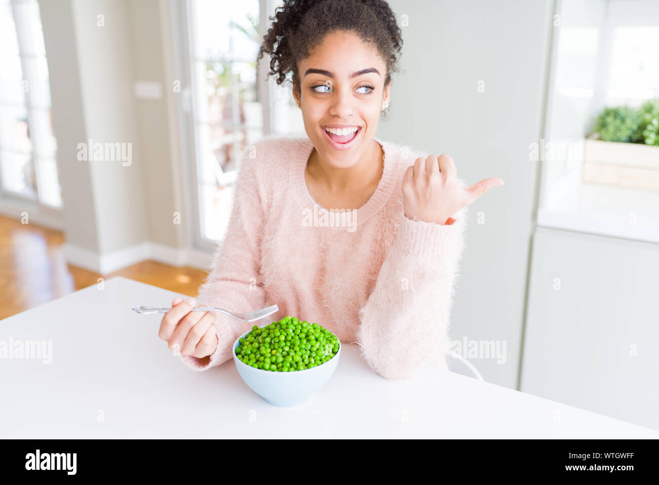 Young african american girl eating healthy green peas pointing and showing with thumb up to the ...