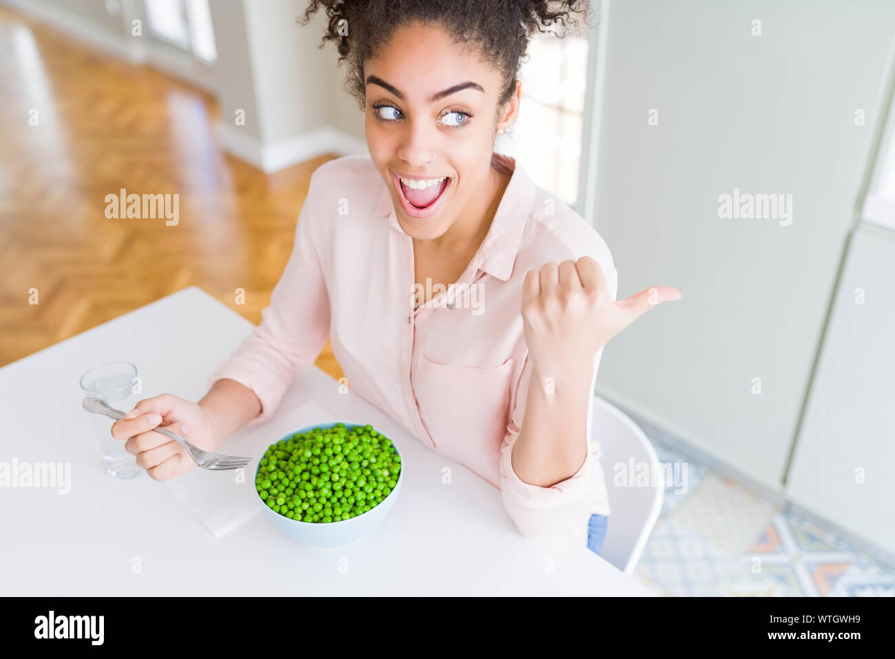 Young african american girl eating healthy green peas pointing and showing with thumb up to the ...